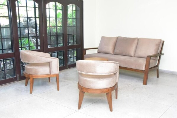 Quarto com sofá bege e duas poltronas combinando, mesa de centro de madeira e uma janela grande com vista para área verde.