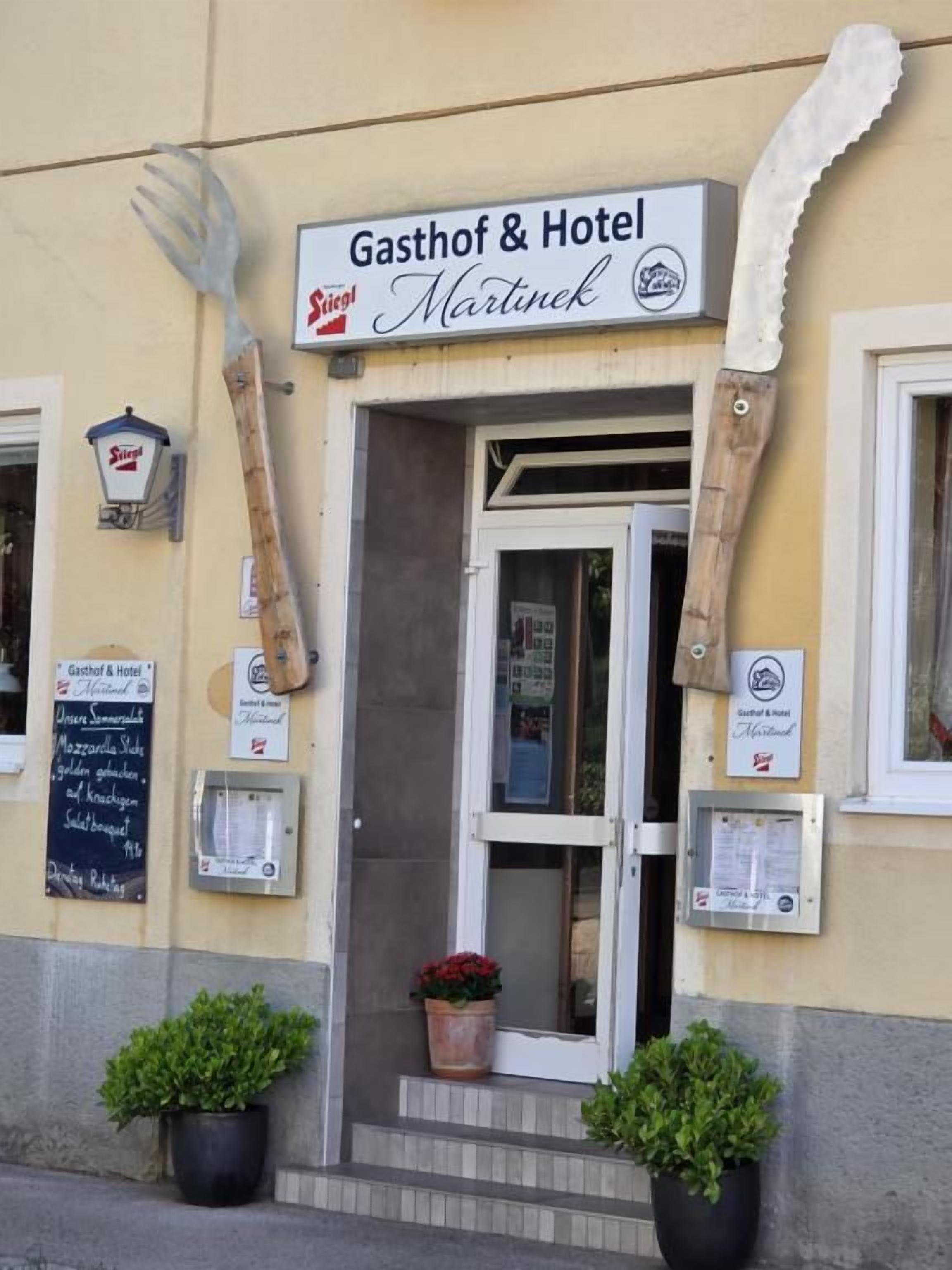 Um edifício com uma placa indicando 'Gasthof & Hotel Martinheck'. Há dois vasos de plantas em frente, uma decoração de espátula de madeira sobre a entrada e um letreiro com texto na parede.