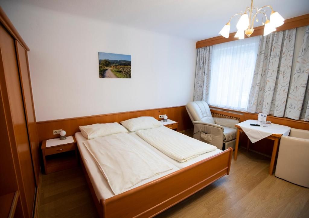 Quarto de hotel com cama, uma cadeira, mesa pequena, criado-mudo e janela com cortinas.
