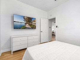 Quarto com cama, cômoda, porta e TV fixada na parede exibindo uma vista panorâmica.