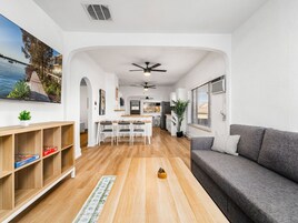 Uma sala de estar moderna com sofá cinza, piso de madeira, área de jantar com mesa e cadeiras, e vista para um corpo de água através de uma janela em arco.