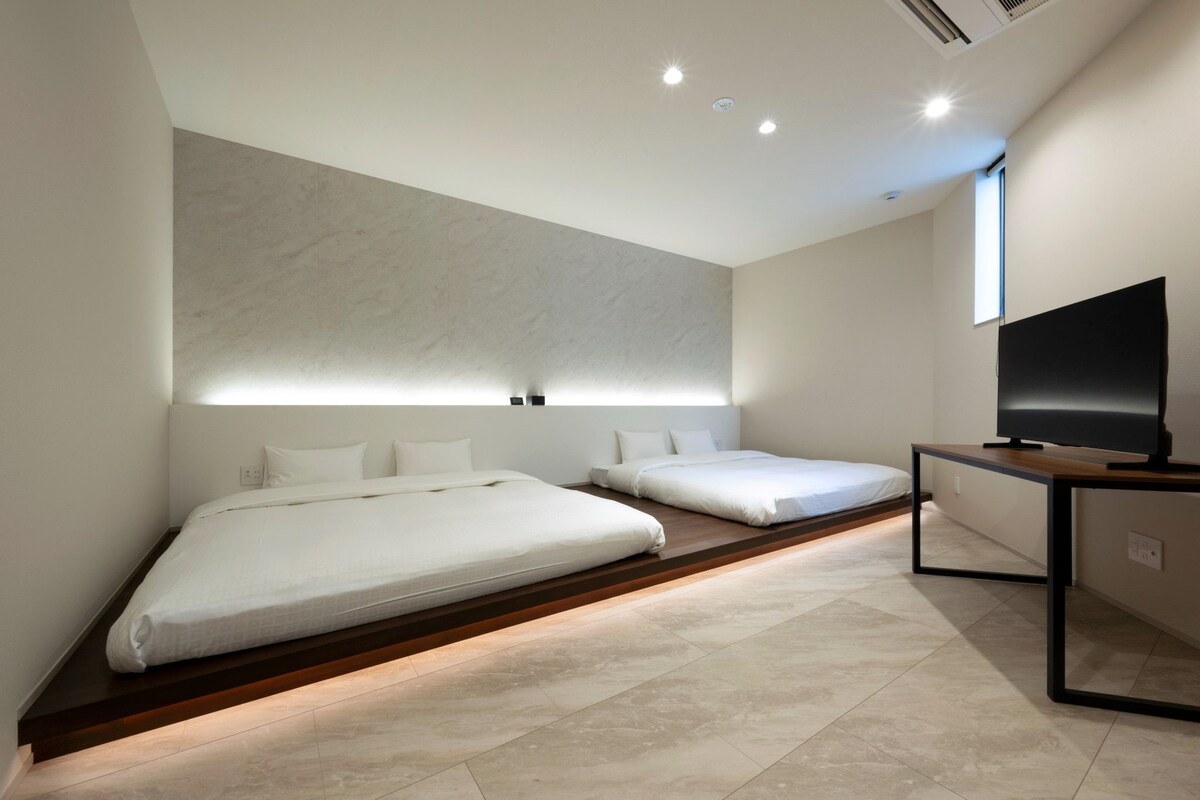 Quarto de hotel moderno com uma cama grande, uma cama menor, uma escrivaninha com televisão de tela plana e iluminação embutida no teto.