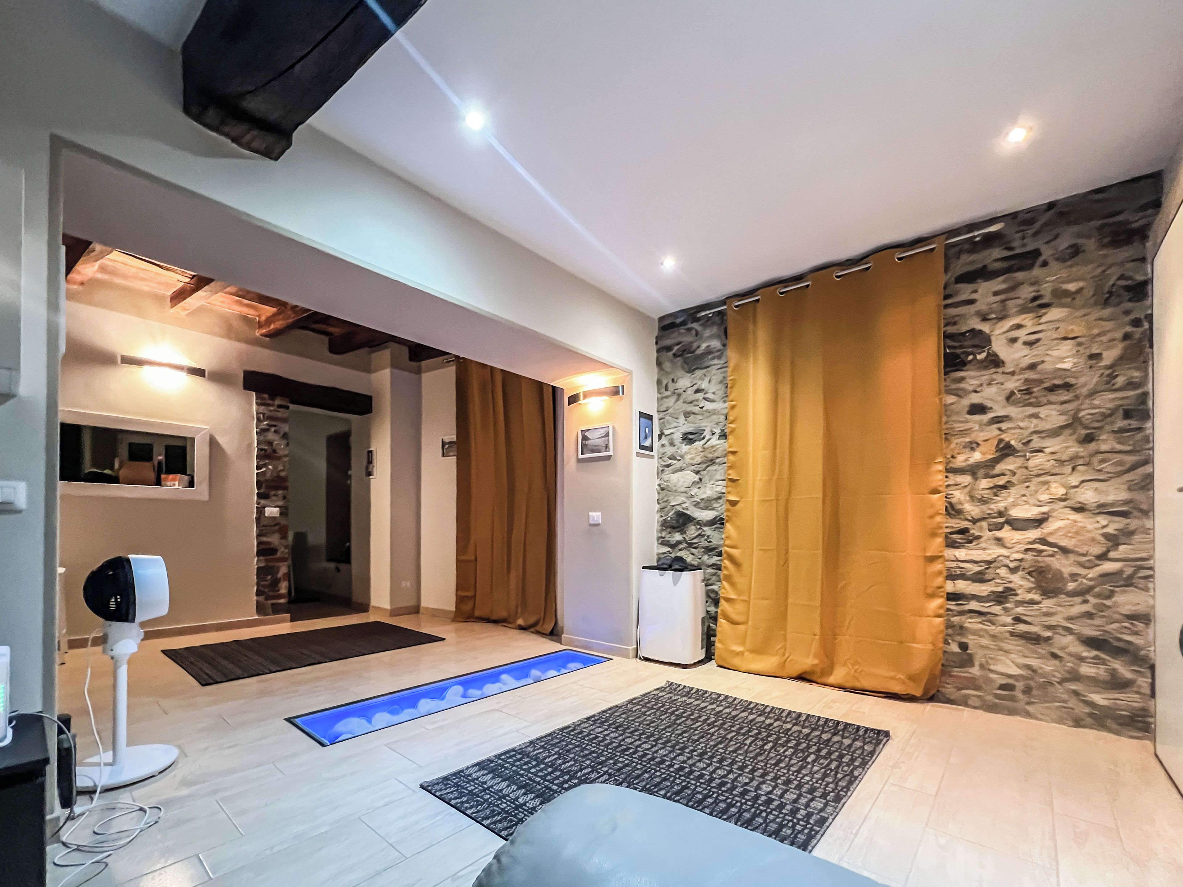 Interior moderno com parede de pedra, caminho iluminado em azul e área de estar com sofá branco.