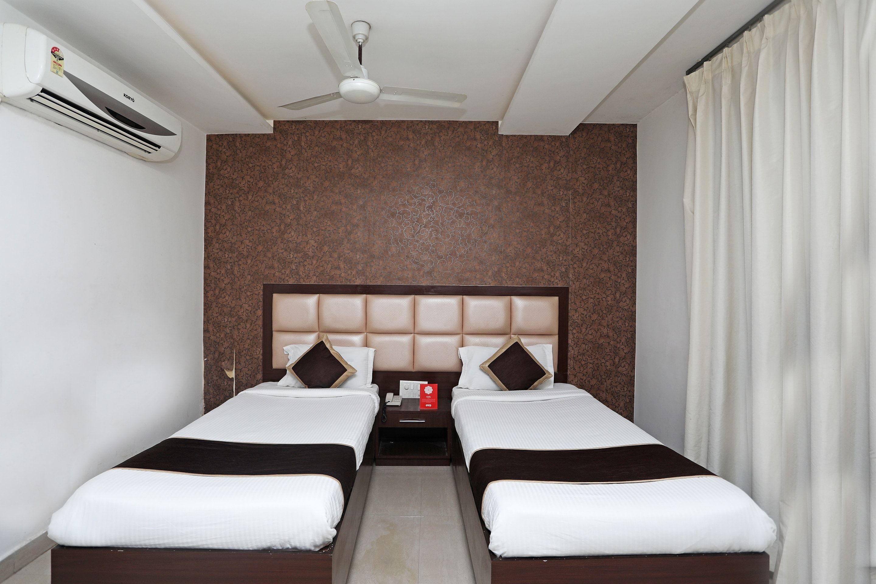 Quarto de hotel com duas camas, ventoinha no teto, aparelho de ar condicionado e cortina na janela.
