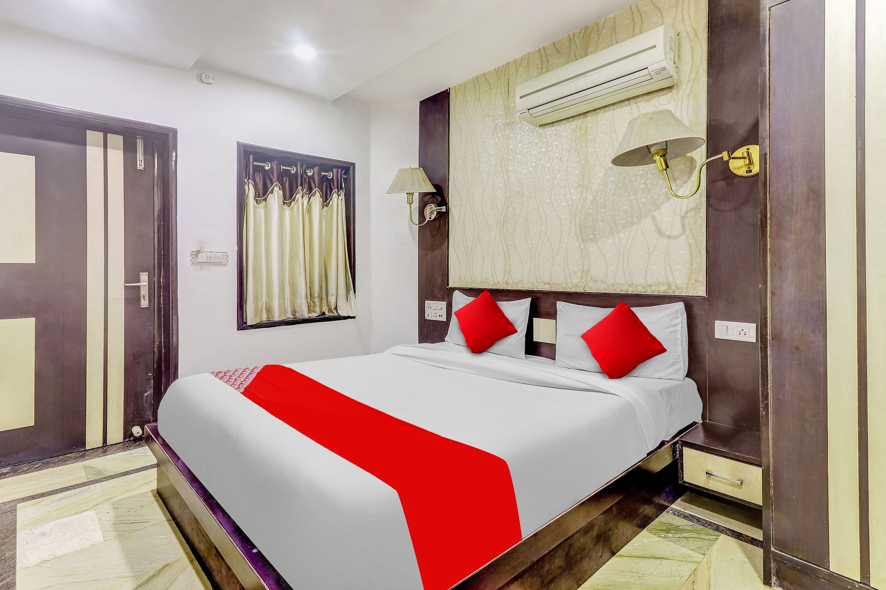 Quarto de hotel com cama, ar condicionado fixado na parede, mesa de cabeceira, luminária e janela com cortinas.