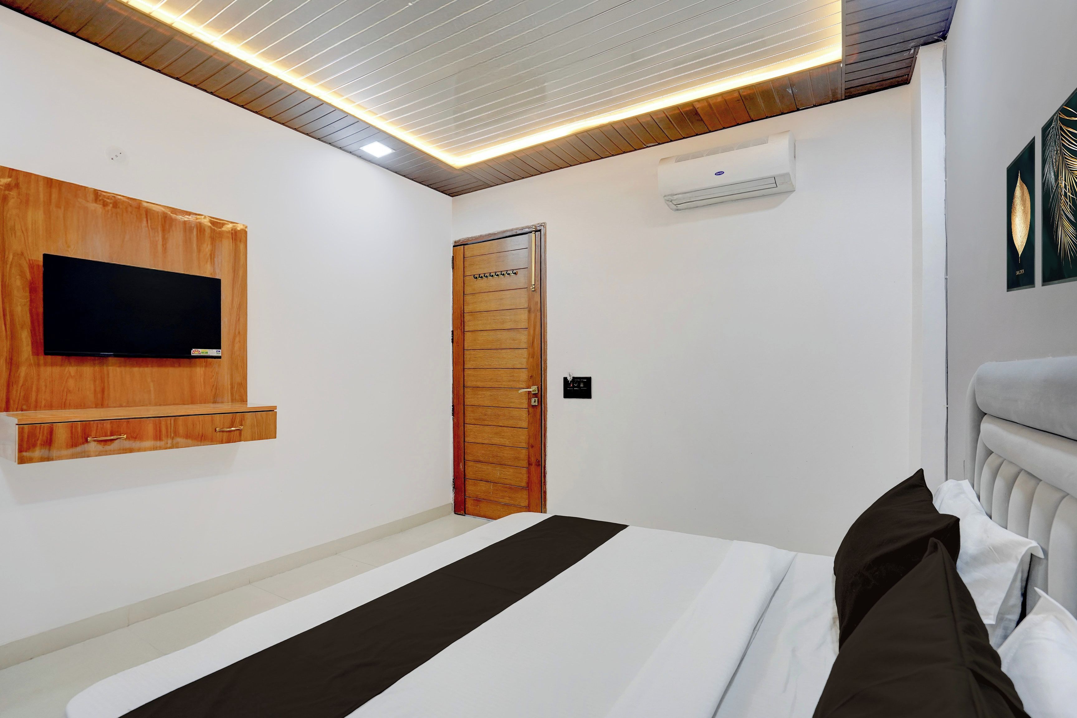Quarto de hotel com cama, televisão fixada em um painel de madeira, aparelho de ar condicionado e uma porta com uma placa.