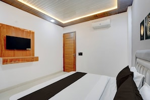 Quarto de hotel com cama, televisão fixada em um painel de madeira, aparelho de ar condicionado e uma porta com uma placa.