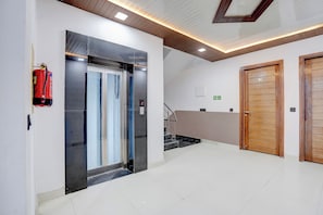 Um elevador moderno com painel de vidro, um extintor fixado na parede e uma escadaria que leva a um andar superior.