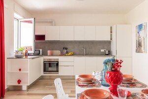 Cozinha moderna com armários brancos, micro-ondas e pia. Há um espaço de jantar com mesa posta para dois, com pratos laranja e um vaso vermelho com flores.