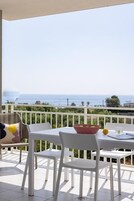 Um pátio externo com mesa e cadeiras, um corrimão branco e vista para o mar.