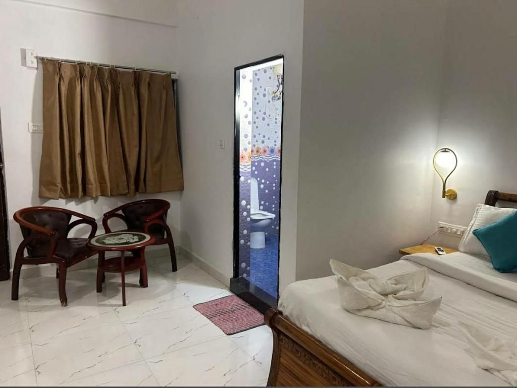 Quarto de hotel com cama, mesa de cabeceira, luminária, janela com cortinas, pequena mesa com cadeiras e vista para o banheiro.
