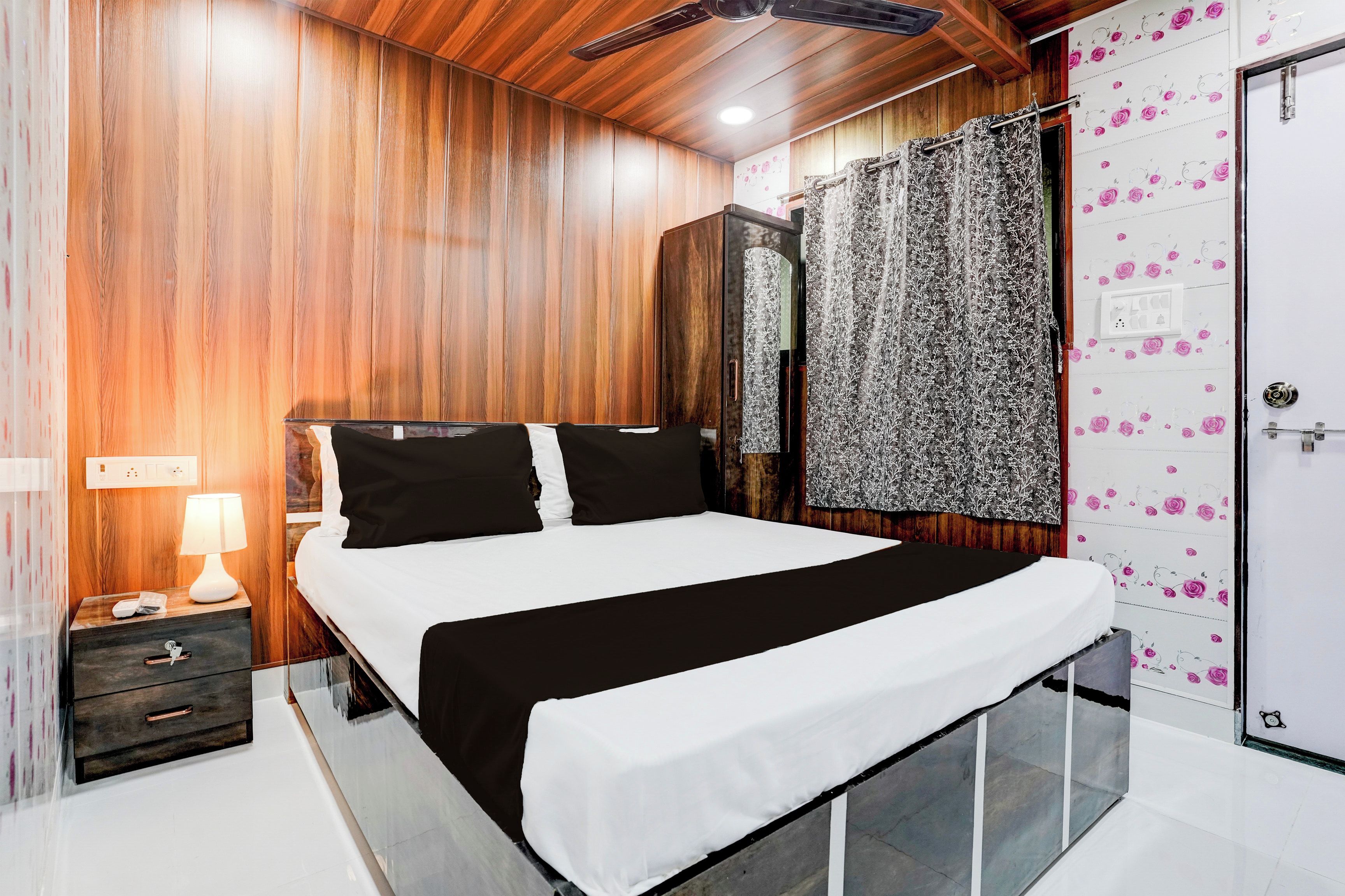 Quarto moderno com parede de madeira, cama com roupa de cama preta e branca, mesa de cabeceira com abajur e porta com papel de parede floral.
