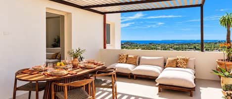Varanda com mesa de jantar posta para quatro pessoas, sofá com almofadas e vista para o mar.