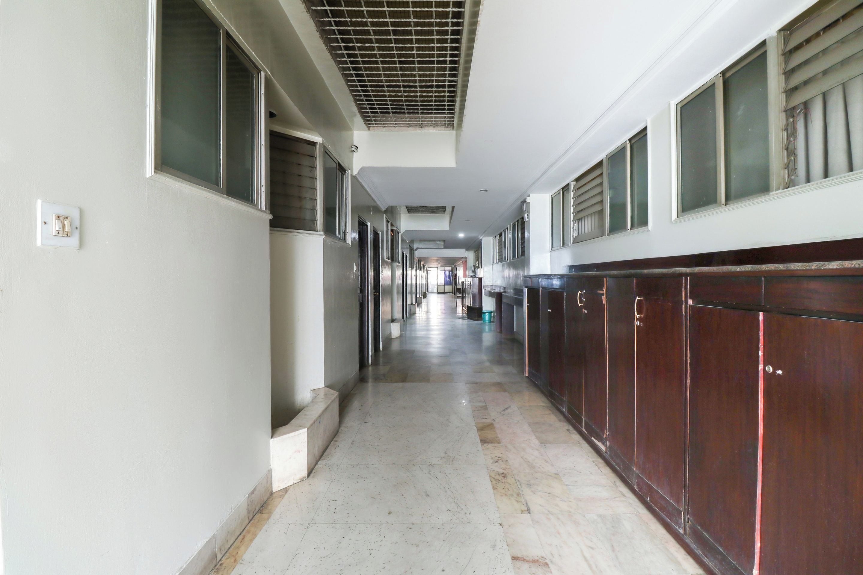 Um corredor comprido e estreito com piso de azulejos, painéis de madeira em uma lateral e uma fileira de janelas ao longo do comprimento.