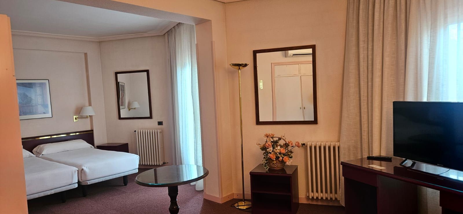 Quarto de hotel com cama, escrivaninha com televisão, mesinha, espelho e radiador.
