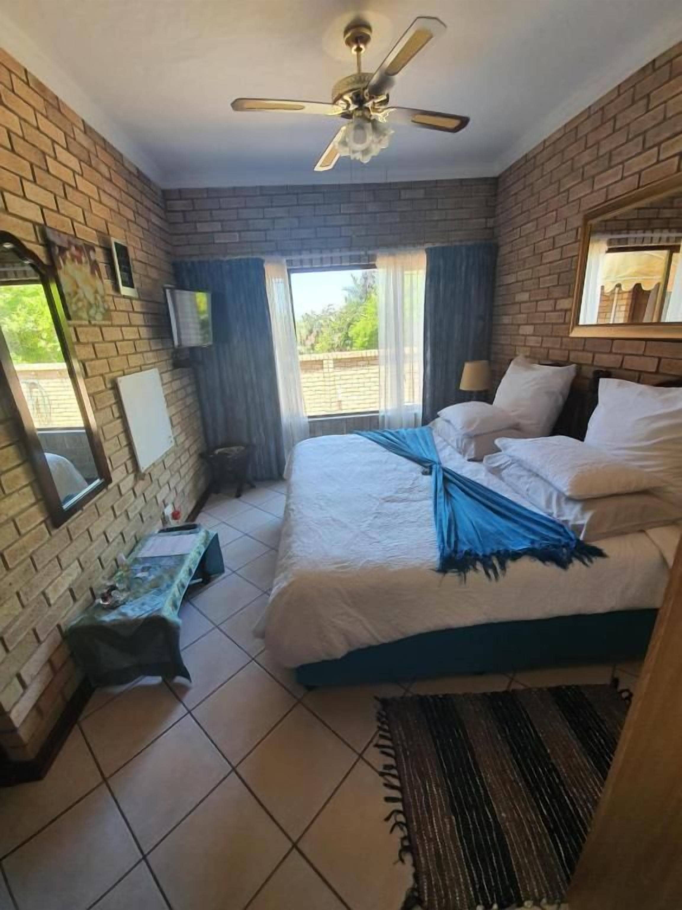 Quarto com cama, ventoinha no teto, janela com cortinas e parede de tijolos.
