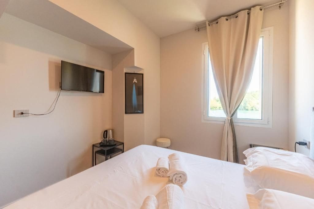 Quarto de hotel com uma cama, televisão na parede, janela com cortinas e uma mesinha com abajur.