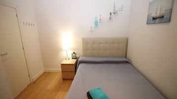 Quarto pequeno e bem organizado, com uma cama de solteiro, mesa de cabeceira com abajur e prateleira na parede com objetos decorativos.