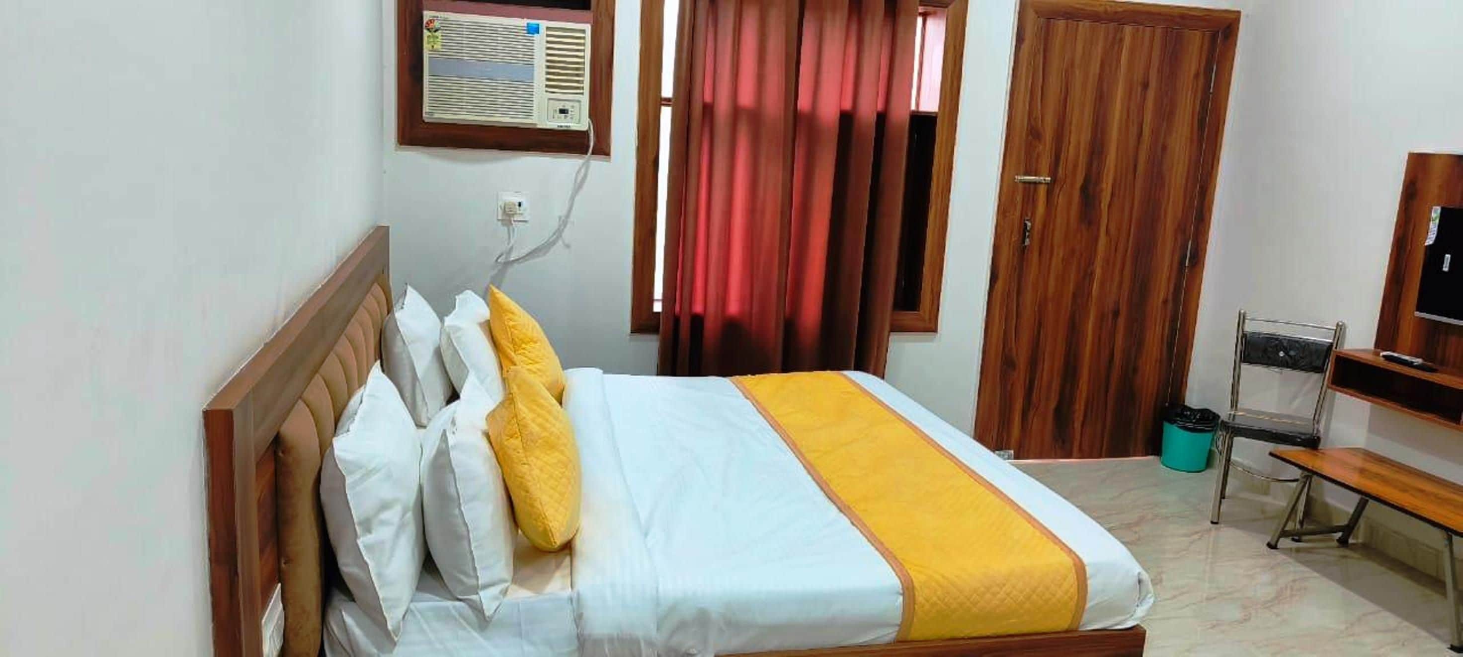 Cama bem arrumada com travesseiros e cobertor, porta de madeira, janela com cortinas e uma pequena lixeira.