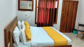 Cama bem arrumada com travesseiros e cobertor, porta de madeira, janela com cortinas e uma pequena lixeira.