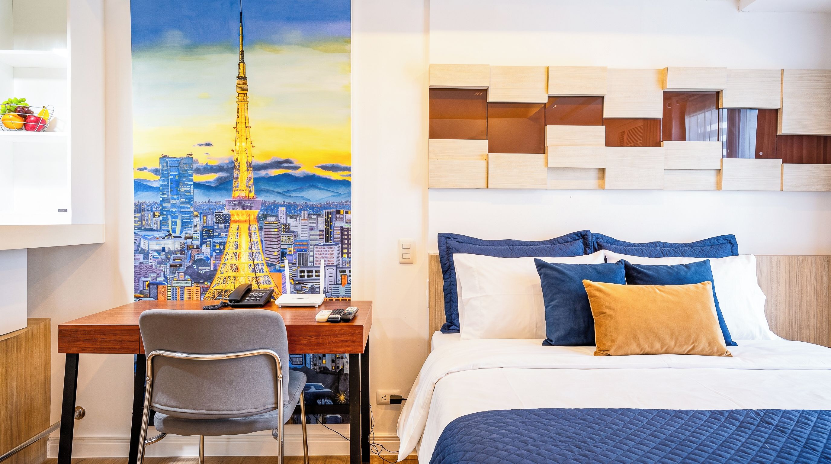 Quarto de hotel com cama, escrivaninha e cadeira. Há um quadro com vista urbana na parede.