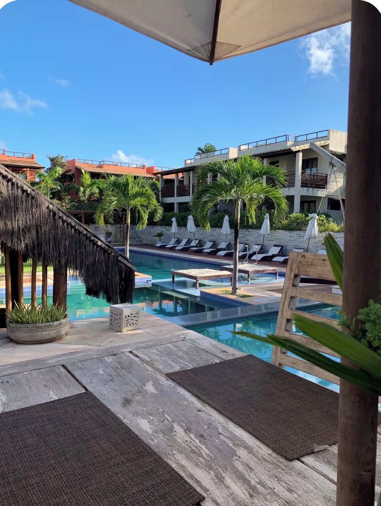 Área de piscina com espreguiçadeiras e guarda-sóis, cercada por edifícios com varandas.