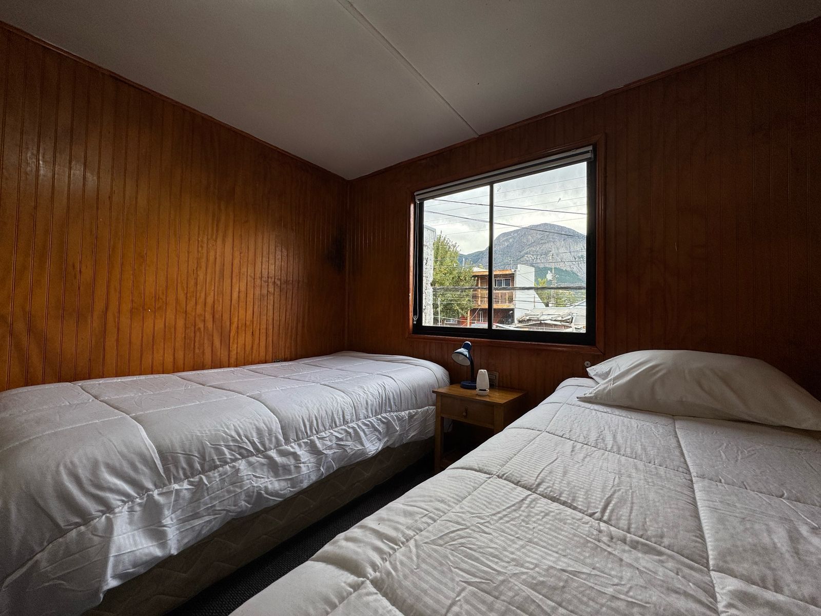 Quarto com duas camas, paredes de madeira, janela com vista para edifícios e montanhas, e mesa de cabeceira com luminária.