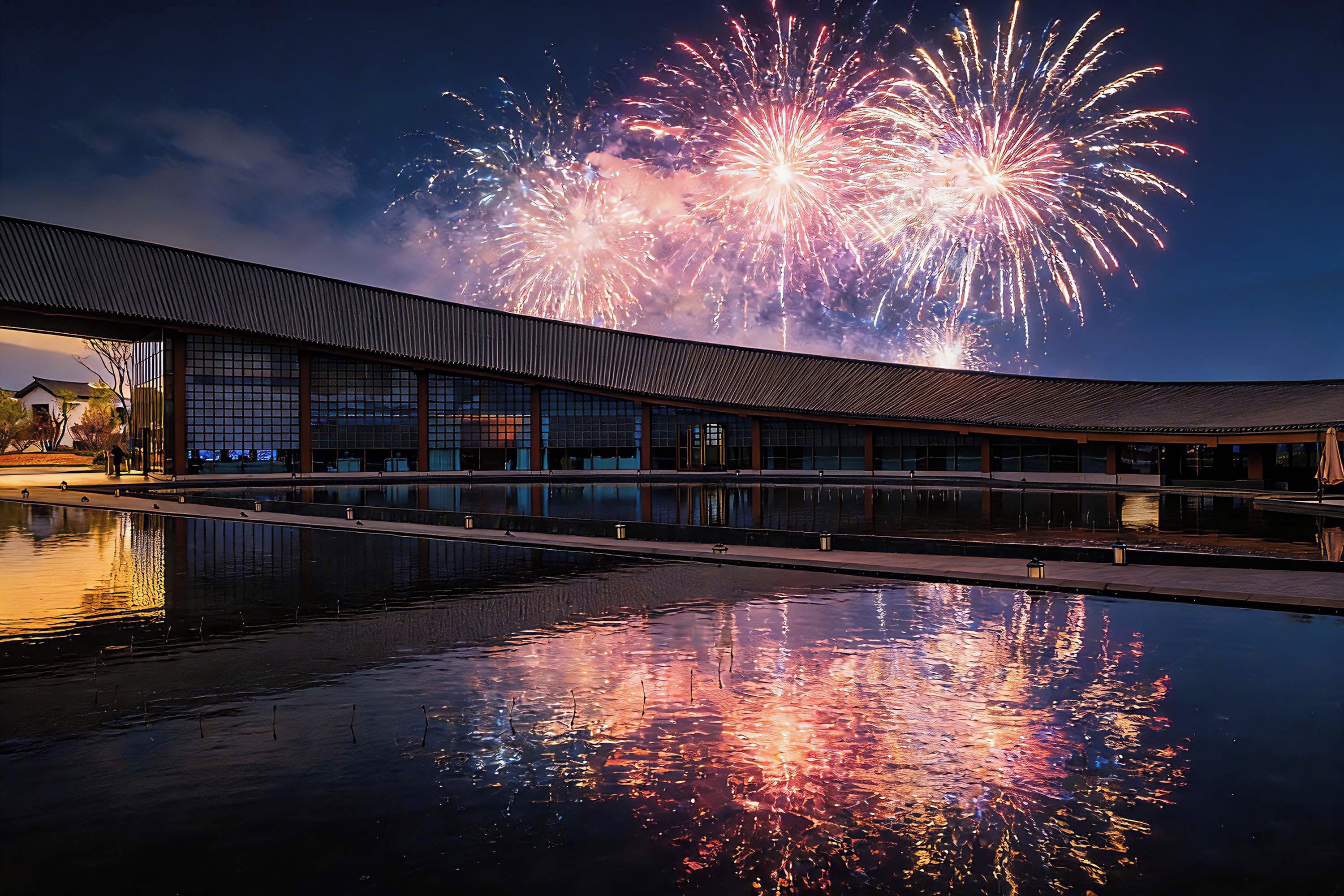 Um edifício com um espelho d'água à frente e fogos de artifício ao fundo.