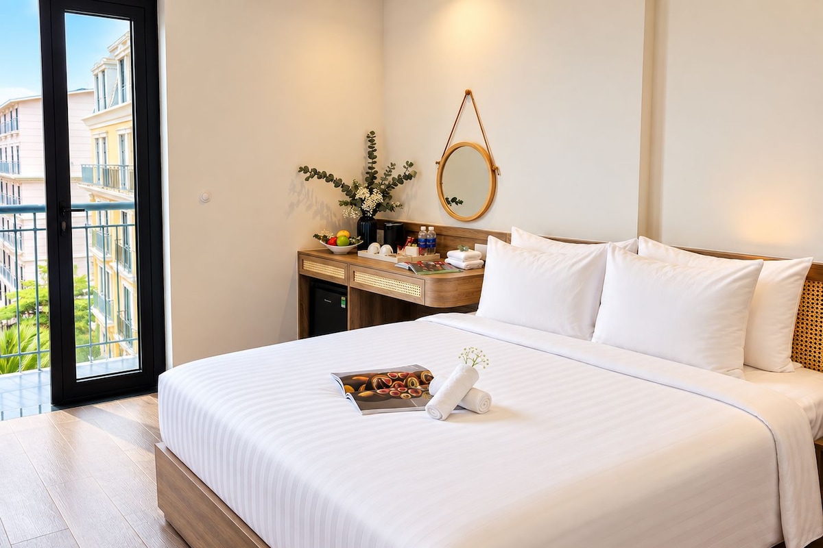 Quarto de hotel com cama grande, mesa de cabeceira, espelho e vista para prédios pela janela.