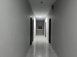 Um corredor com paredes brancas e chão de azulejos, que leva a um quarto com janela e porta.