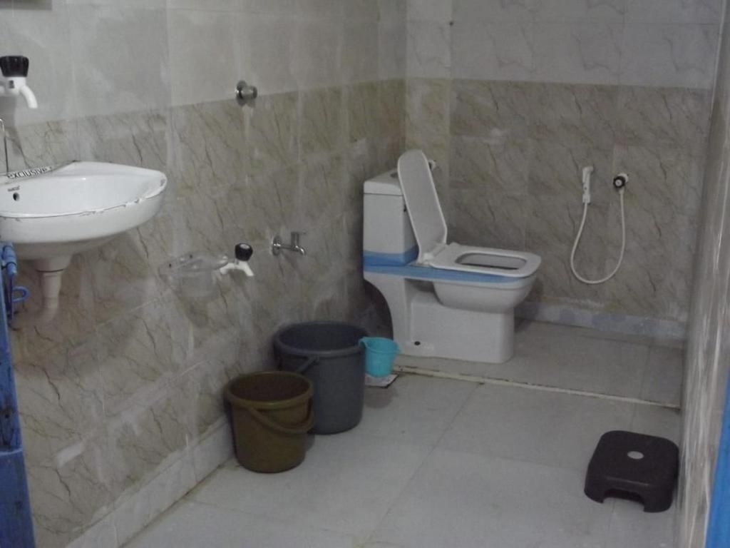 Casa de banho com lavatório, sanita e chuveiro. Inclui contentores de lixo e um banquinho.