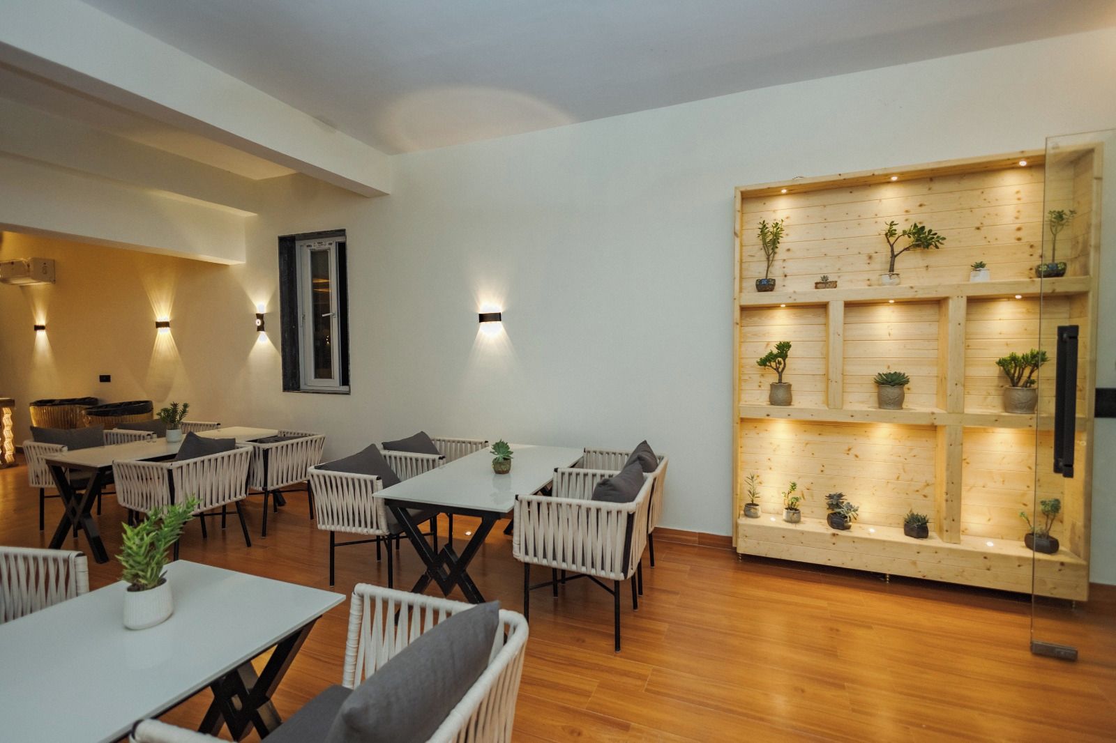 Uma área de refeições moderna, com mobiliário de vime branco, piso de madeira e um aparador de plantas na parede.