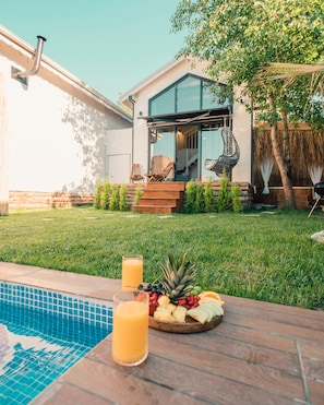 Um quintal com piscina, deck de madeira, mesa de jantar com frutas e sucos, e uma casa com porta de vidro e área de estar externa.