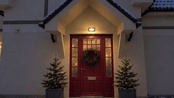 Uma casa com uma porta vermelha, duas árvores de Natal em vasos e um chão coberto de neve.