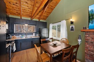 Cozinha compacta com vigas de madeira no teto, área de refeições com mesa e cadeiras, e janela com vista para uma montanha.