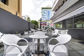 Uma área externa com assentos de vime e mesas, vista da rua com edifícios, e um céu claro.