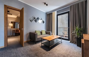 Quarto de hotel moderno com sofá, mesa de centro de madeira, escrivaninha e uma janela grande com cortinas.