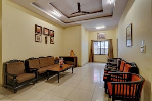 Uma sala de estar ampla com um conjunto de sofás, uma mesa de centro e uma mesa de jantar. Há quadros nas paredes, um ventilador de teto e uma janela com cortinas.