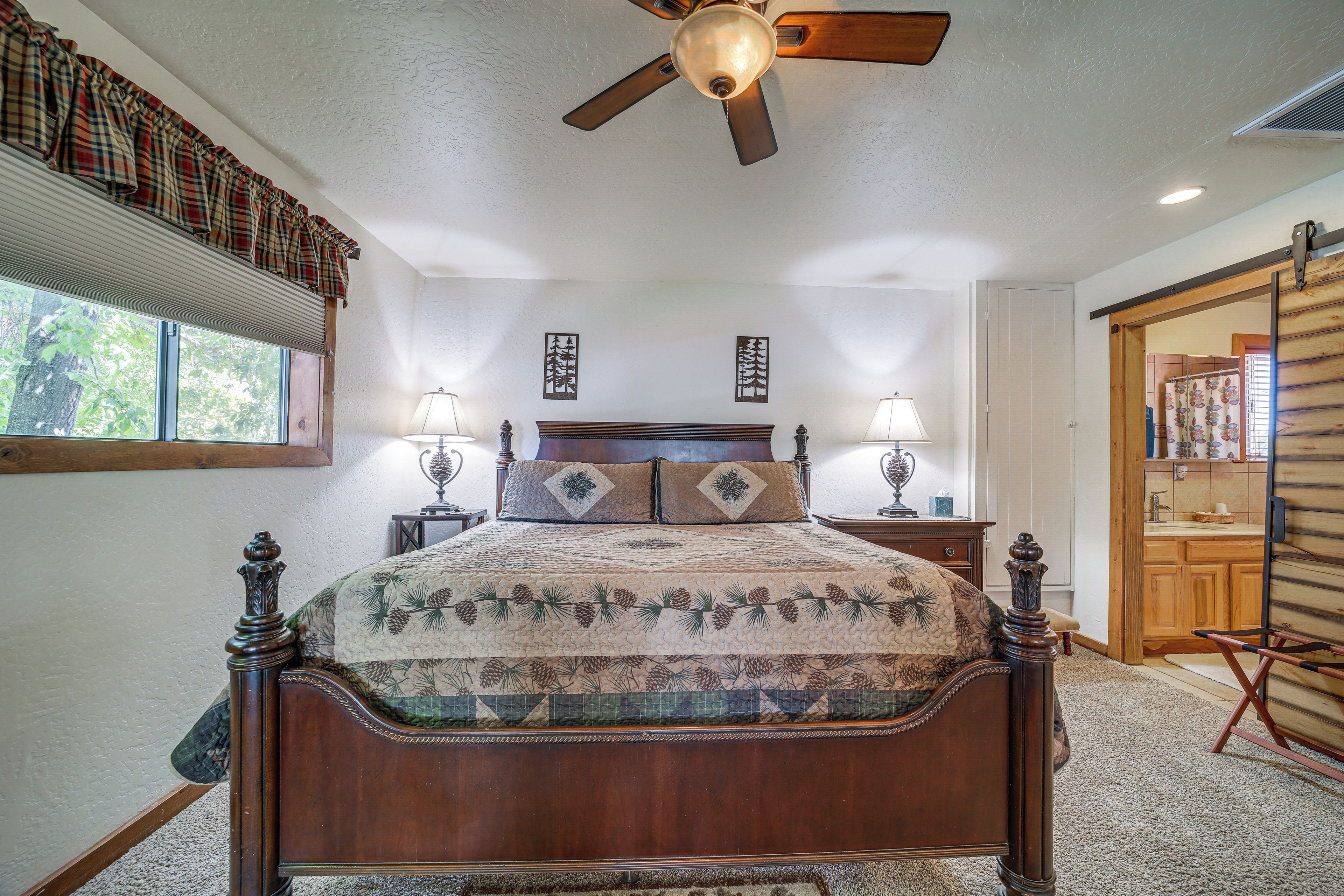 Quarto com cama de madeira, duas luminárias de cabeceira, ventilador de teto e vista para o exterior através de uma janela com persianas.