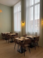 Área de refeições com mesas e cadeiras, grandes janelas com cortinas e uma parede de tijolos visível do lado de fora.
