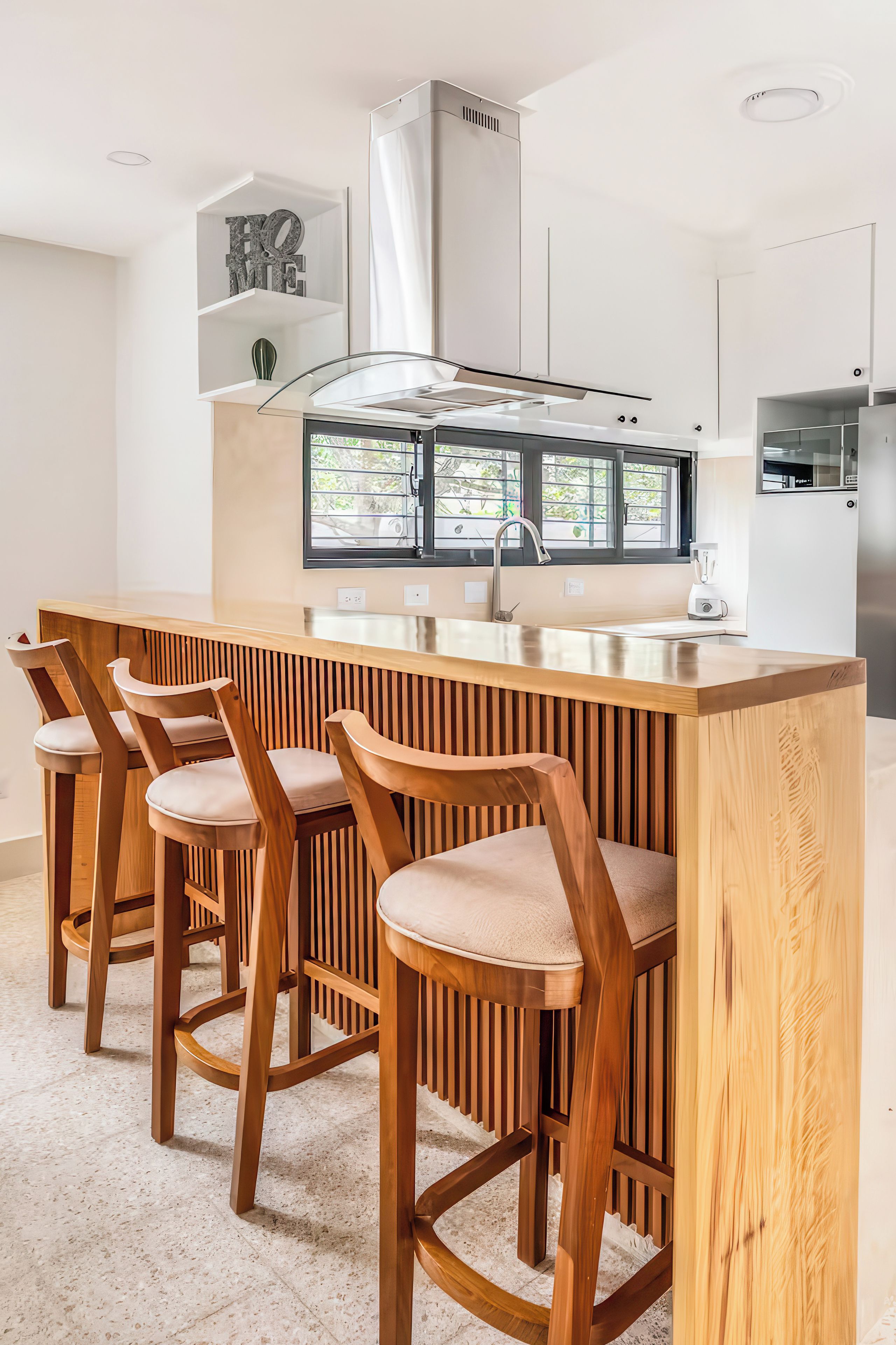 Cozinha moderna com ilha central, banquetas de bar e janela com vista para o verde.