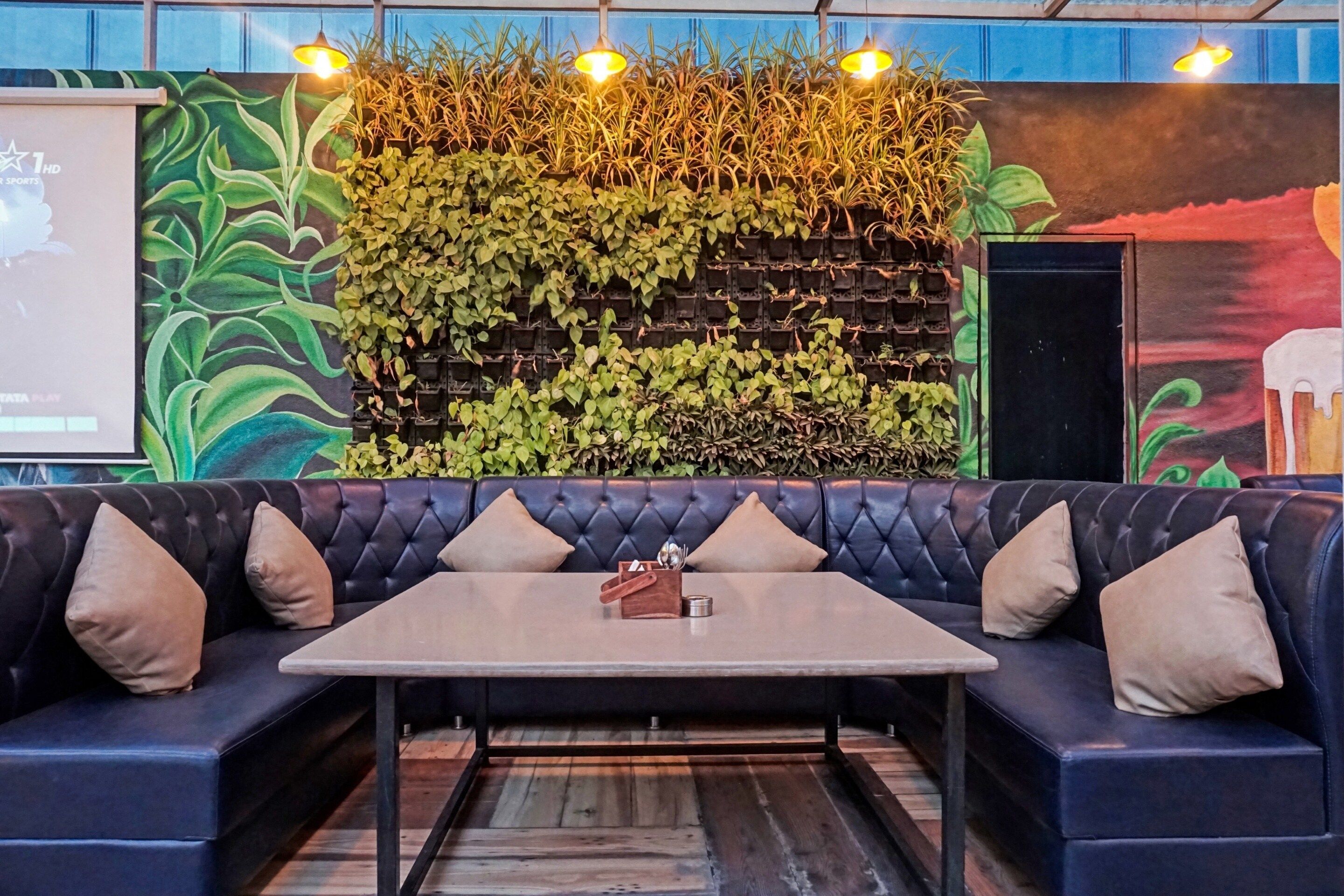 Uma área de estar com mesa e bancos acolchoados, parede com jardim vertical e tela de projeção.