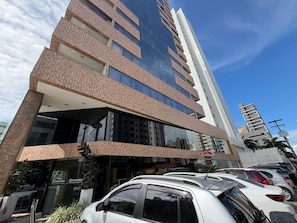 Um hotel com vários andares, fachada moderna, grandes janelas e estacionamento com diversos carros.