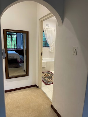 Um corredor com um espelho refletindo um quarto, um banheiro com banheira e um tapete estampado no chão.
