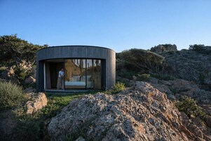 Um edifício circular com fachada de vidro, uma cama no interior e uma pessoa ao lado da entrada.