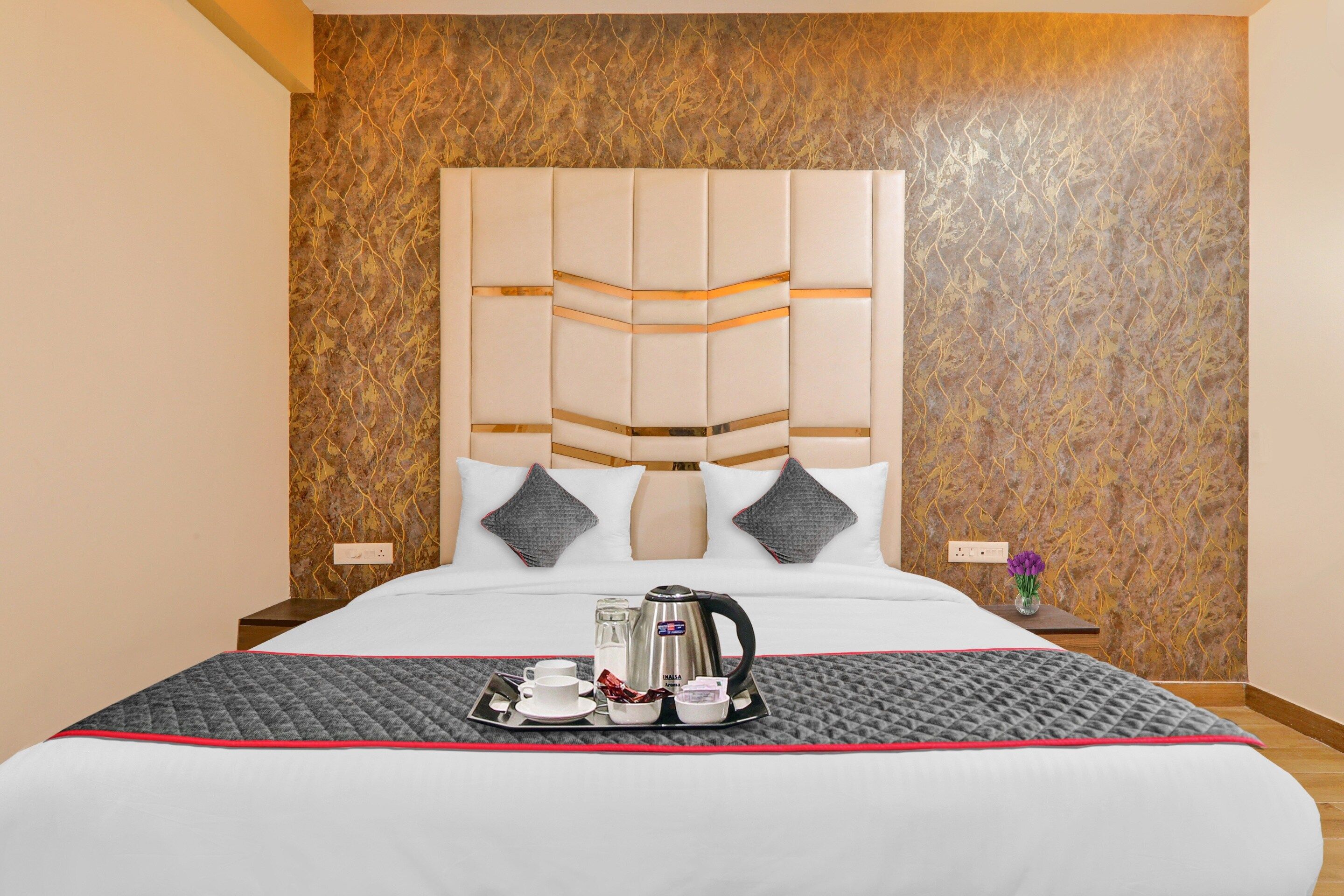 Quarto de hotel com uma cama grande, cabeceira com listras horizontais, bandeja com chaleira e xícaras, e um vaso com flores na mesa de cabeceira.
