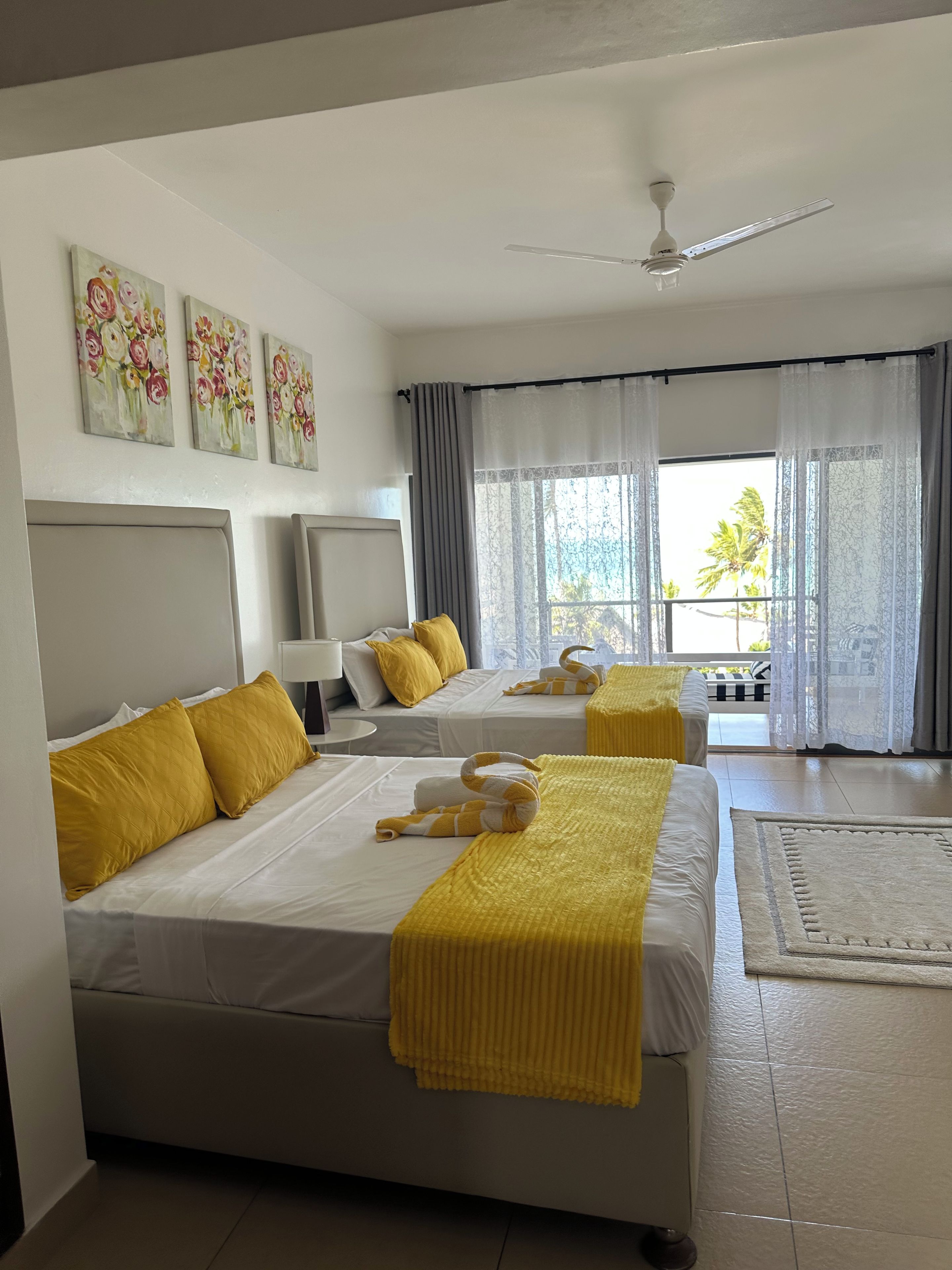 Quarto de hotel com duas camas, ventilador de teto e vista para palmeiras.