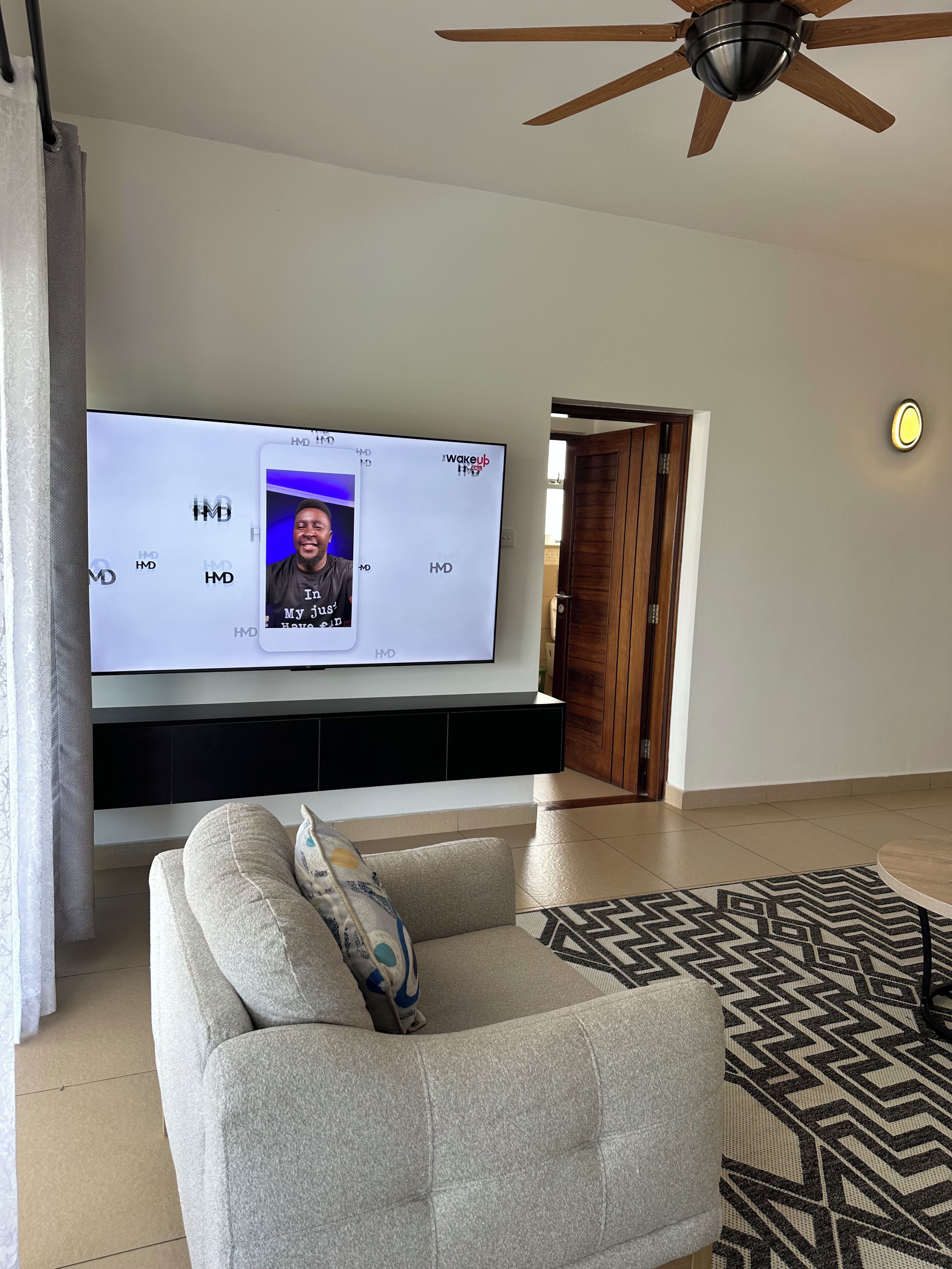 Sala de estar com televisão de tela plana na parede, ventilador de teto, sofá e tapete estampado.
