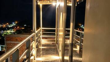 Um corredor com corrimão de metal, porta de vidro e vista para paisagem urbana à noite.