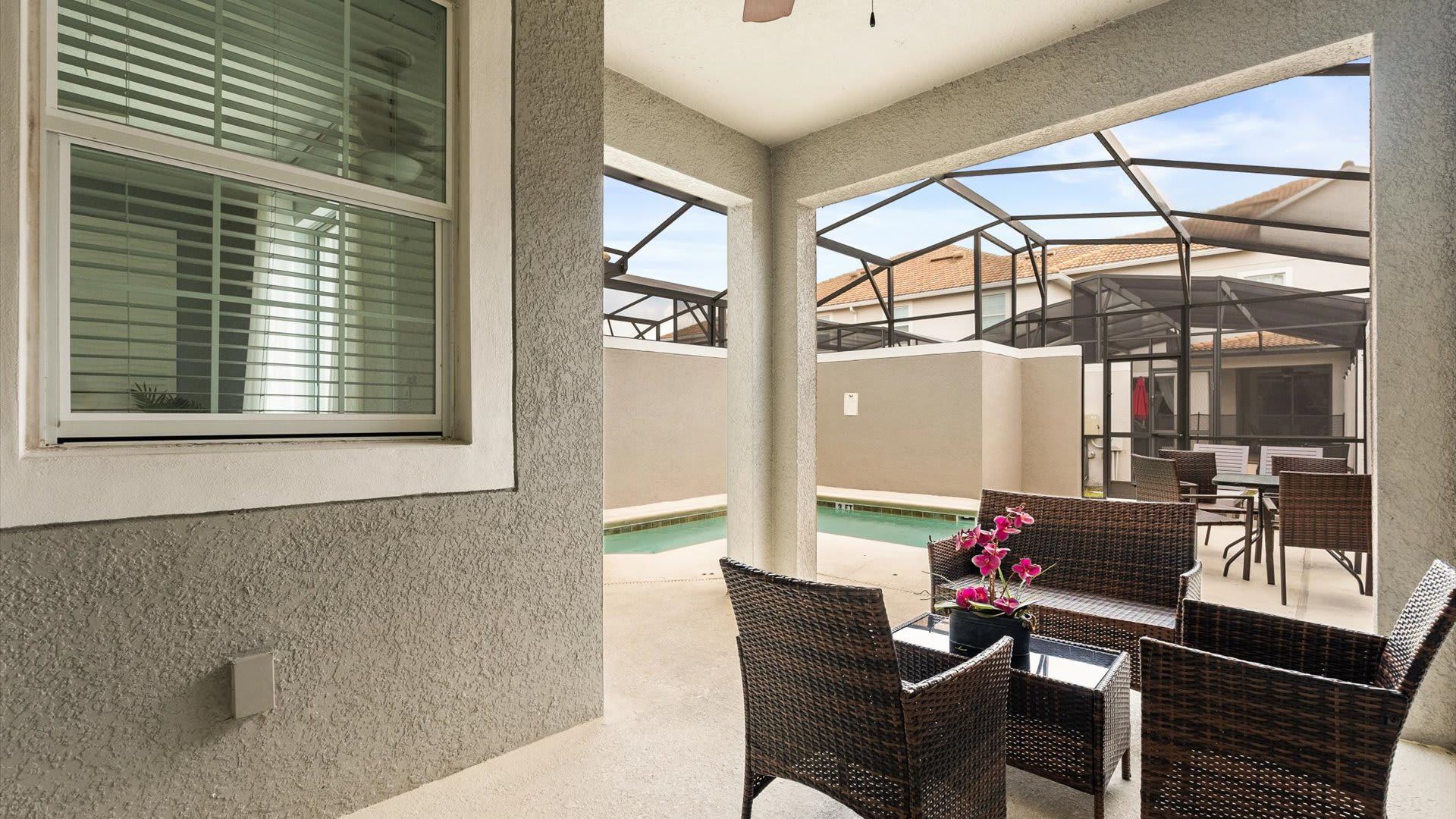 Uma varanda coberta com móveis de vime, uma mesa de vidro e vista para uma piscina.